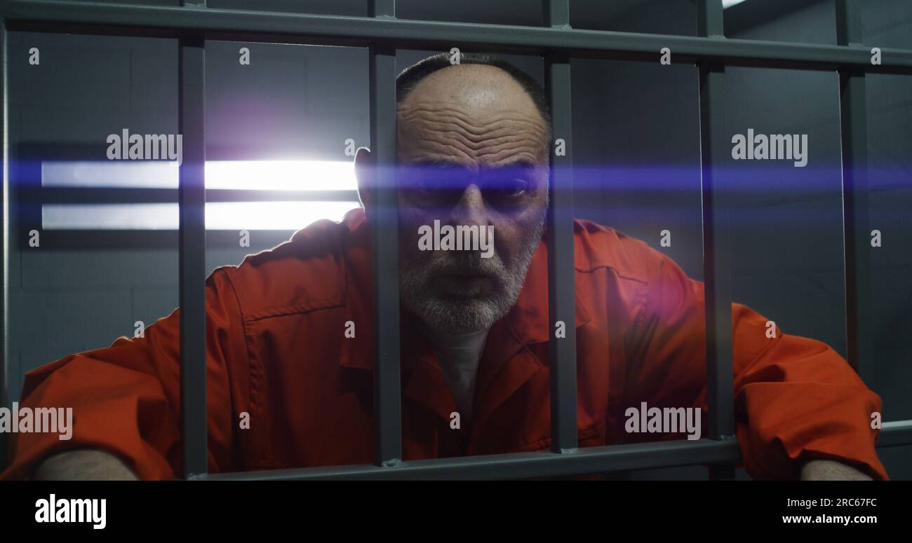 Elderly prisoner in orange uniform holds hands on metal bars, looks at ...