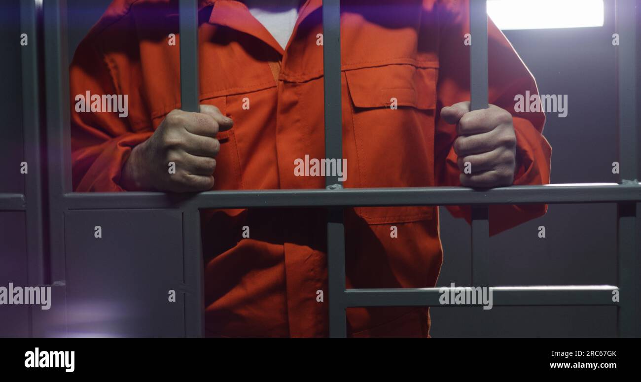 Elderly prisoner in orange uniform holds metal bars, stands in prison ...
