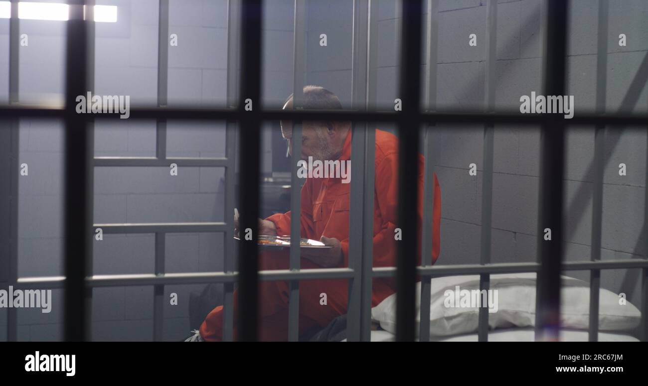 Elderly prisoner in orange uniform sits in prison cell, eats food ...