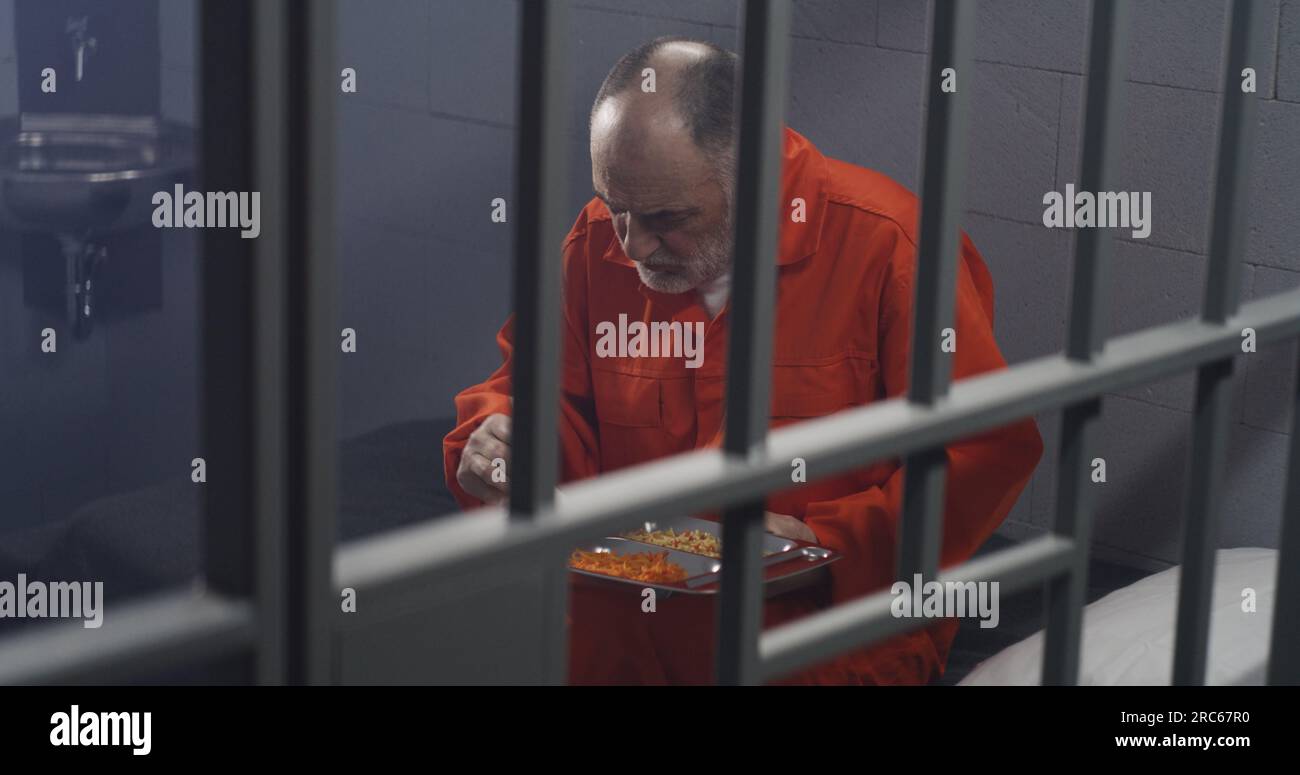 Elderly prisoner in orange uniform sits in prison cell, eats food ...