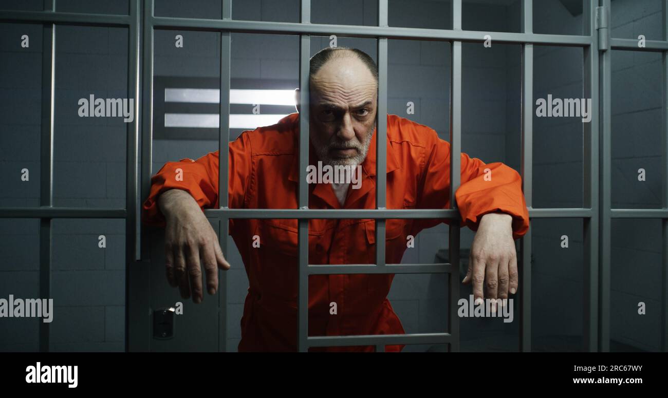 Elderly prisoner in orange uniform holds hands on metal bars, looks at ...