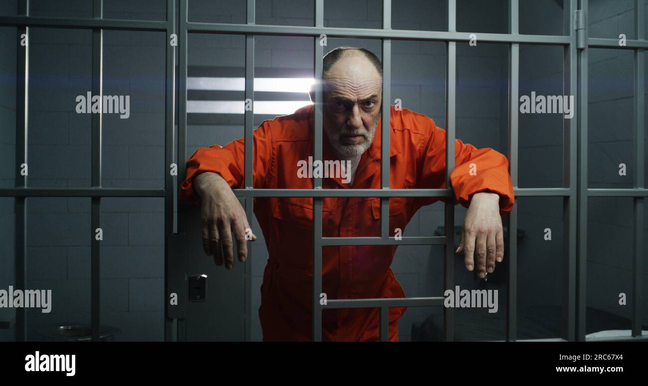 Elderly prisoner in orange uniform holds hands on metal bars, looks at ...