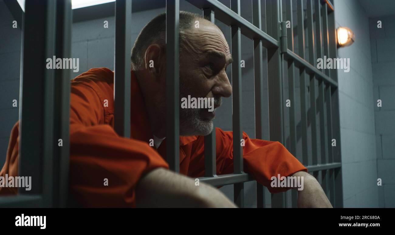 Elderly prisoner in orange uniform stretches his hands, leans on metal ...