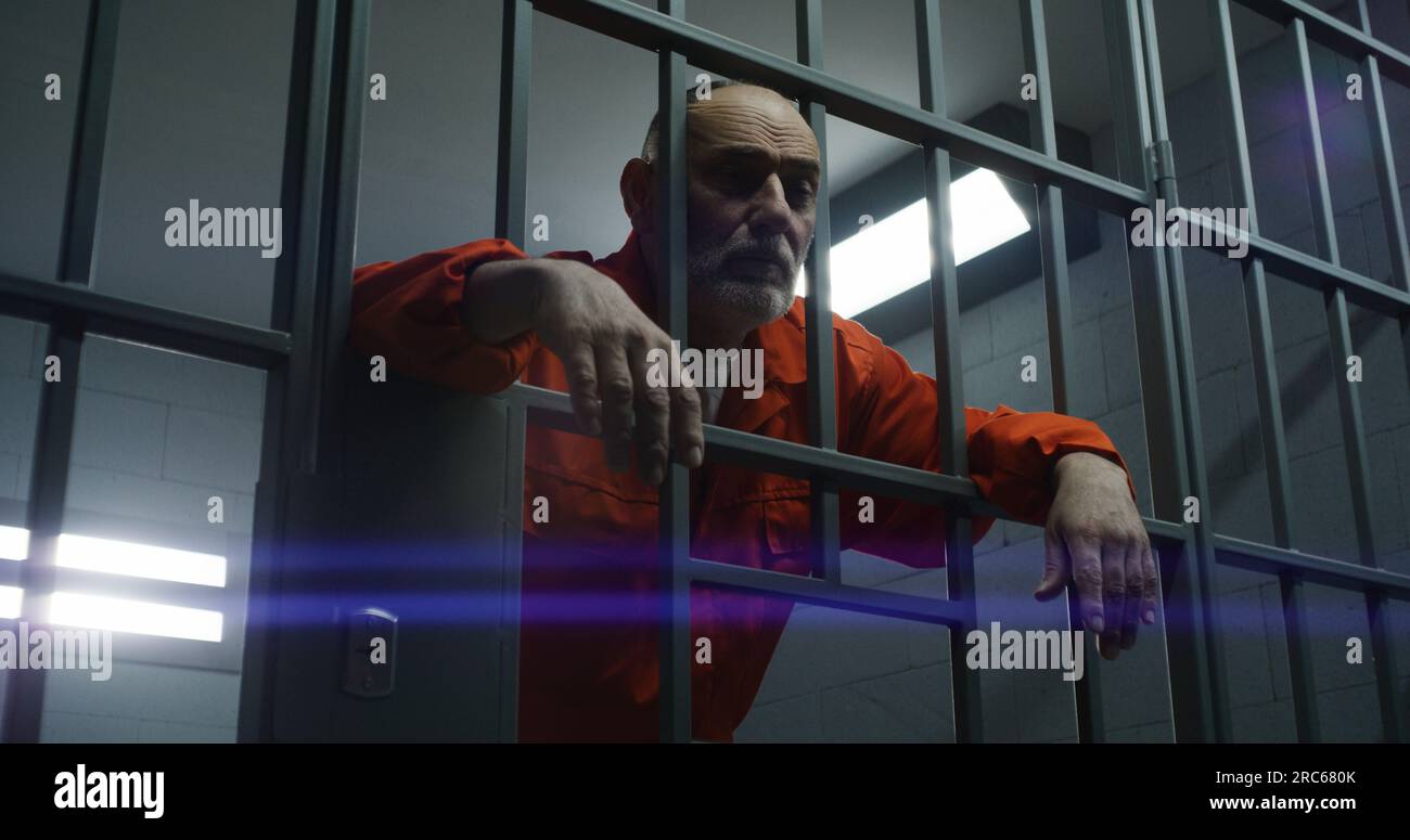 Elderly prisoner in orange uniform holds hands on metal bars in jail ...