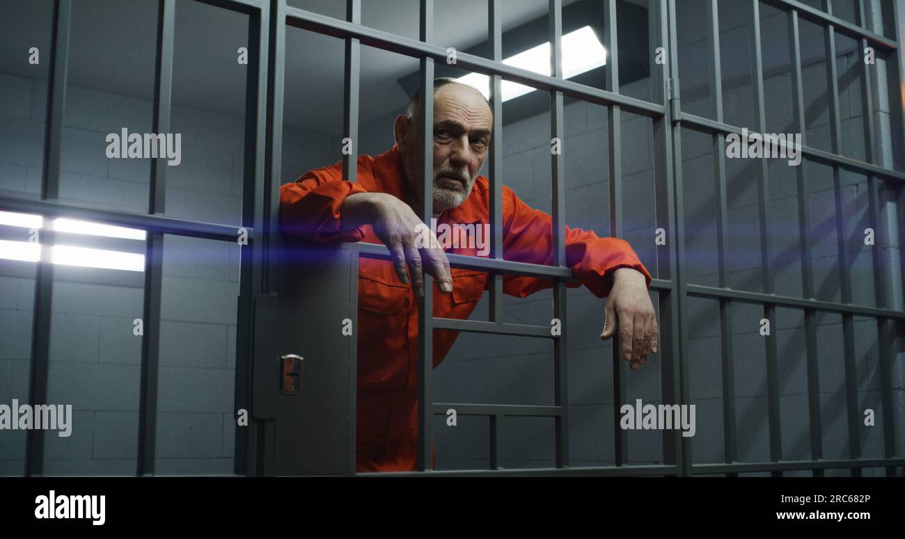 Elderly prisoner in orange uniform holds hands on metal bars in jail ...