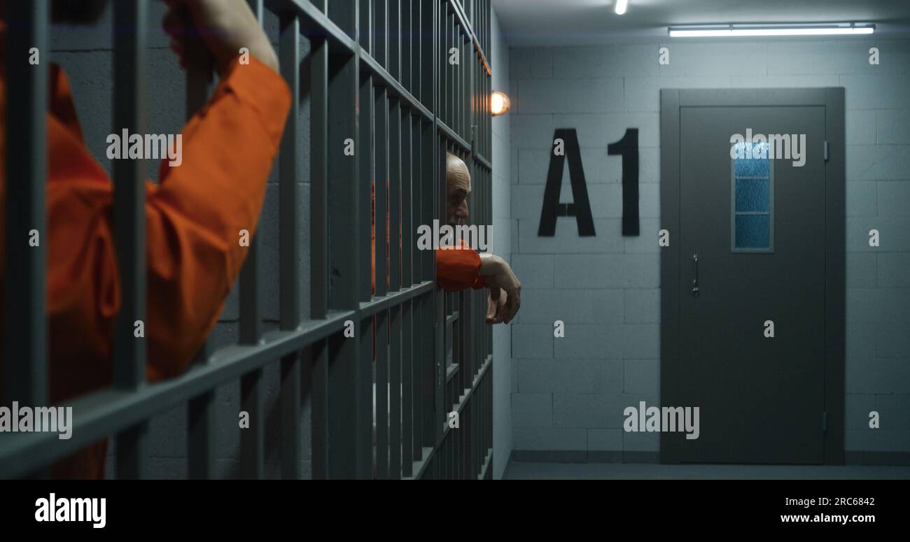 Elderly prisoner in orange uniform stretches fingers, leans on metal ...