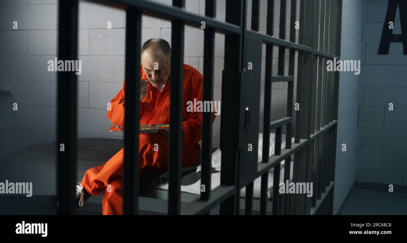Elderly prisoner, inmate in orange uniform eats food sitting in prison ...