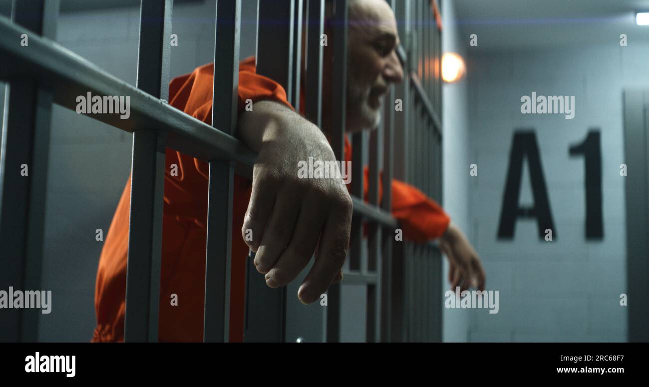 Elderly prisoner in orange uniform stretches his hands, leans on metal ...