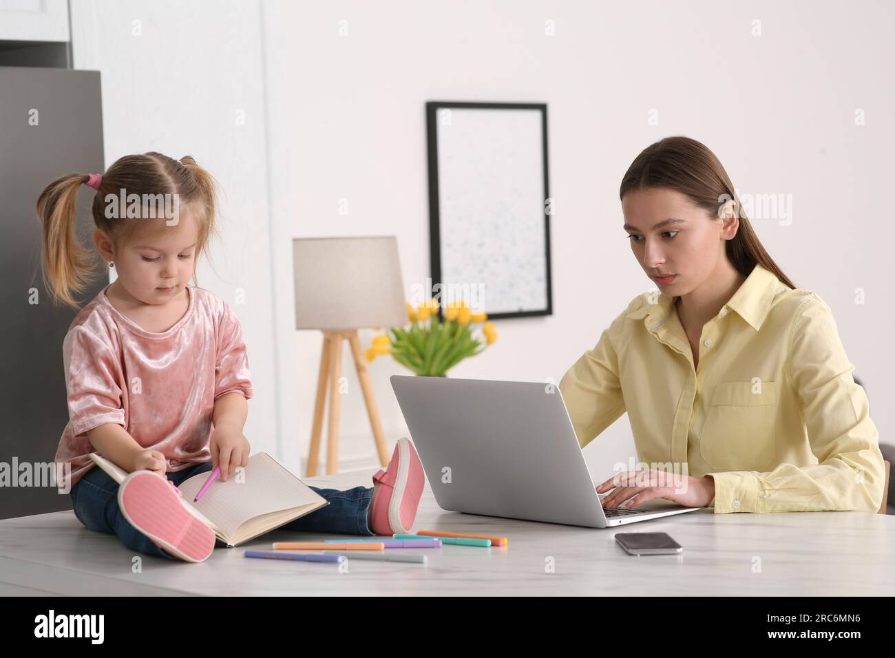 Mother with laptop working remotely while her daughter drawing at home ...