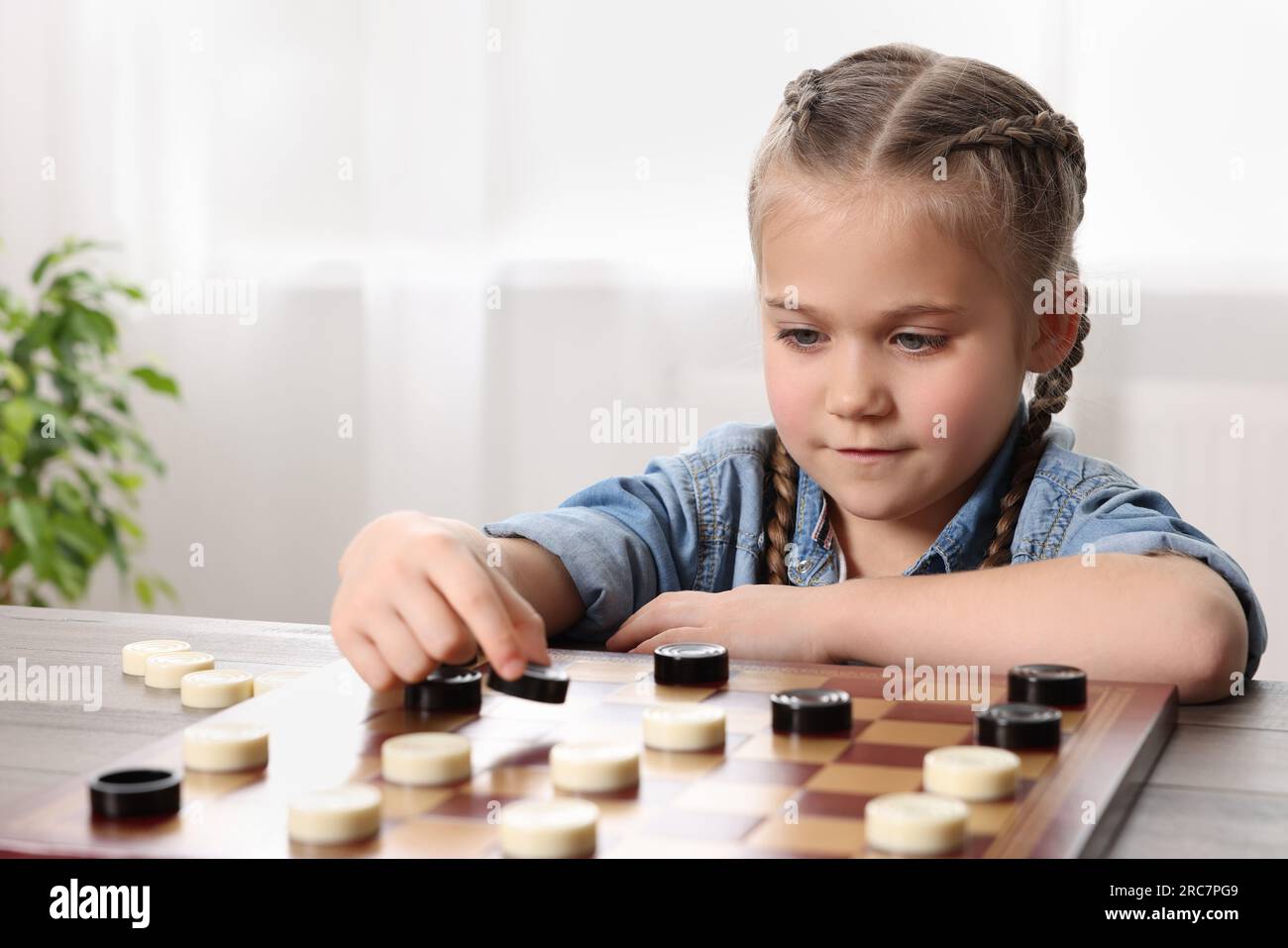 Playing checkers. Little girl thinking about next move at table in room ...