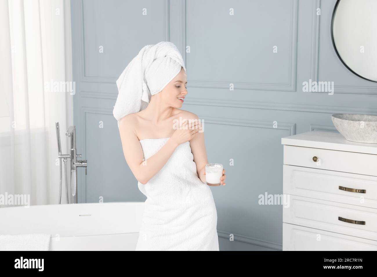 Beautiful young woman applying body cream onto shoulder in bathroom ...