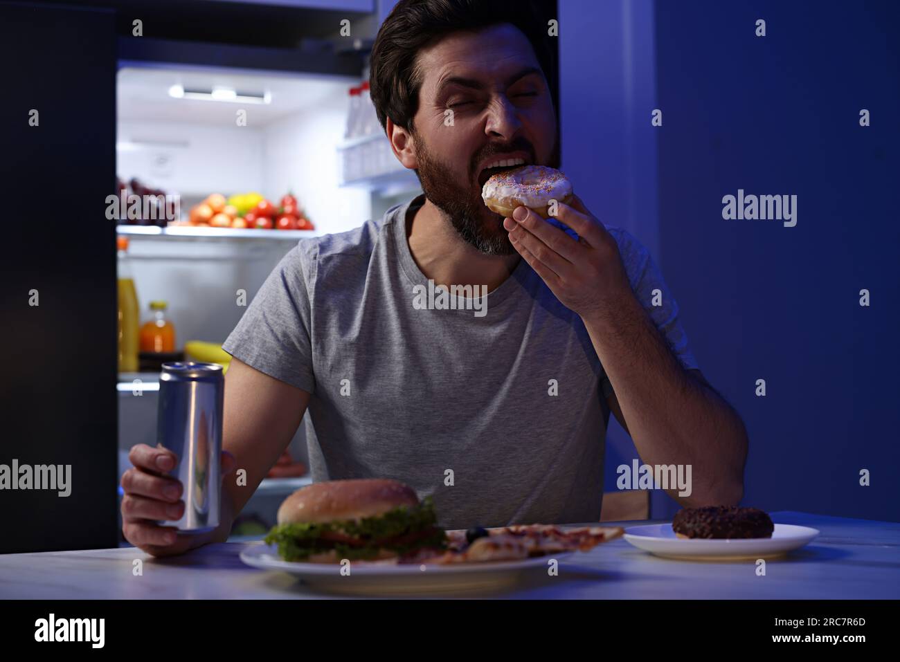 Man holding tin can and eating donut in kitchen at night. Bad habit ...