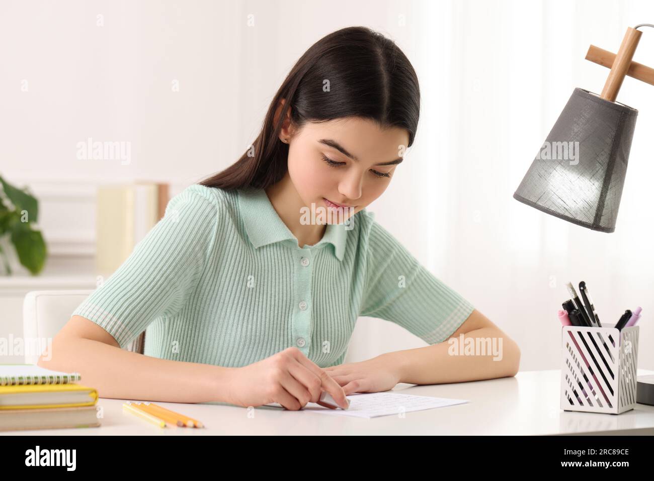Teenage girl erasing mistake in her notebook at white desk indoors ...