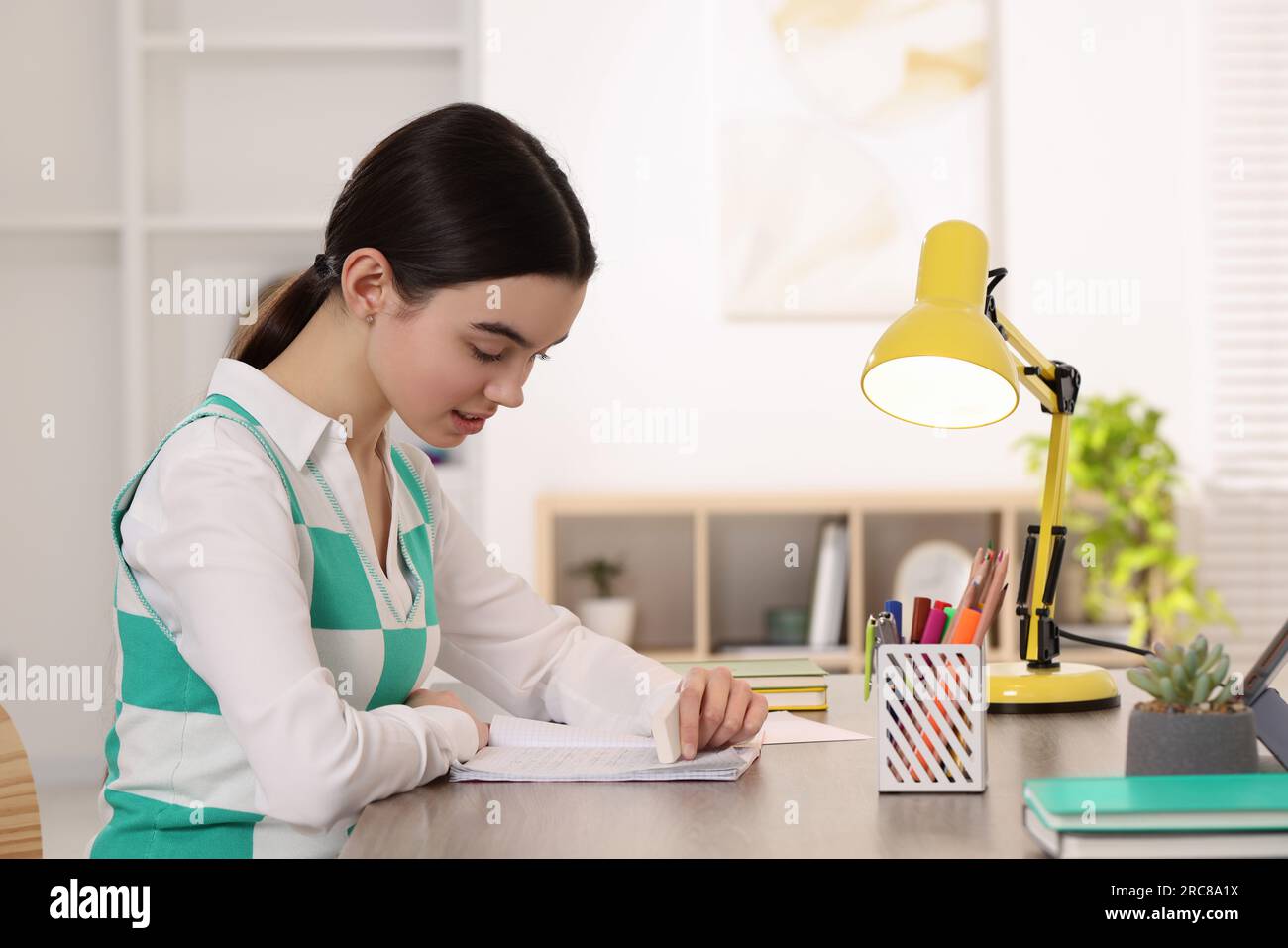 Teenage girl erasing mistake in her notebook at wooden desk indoors ...