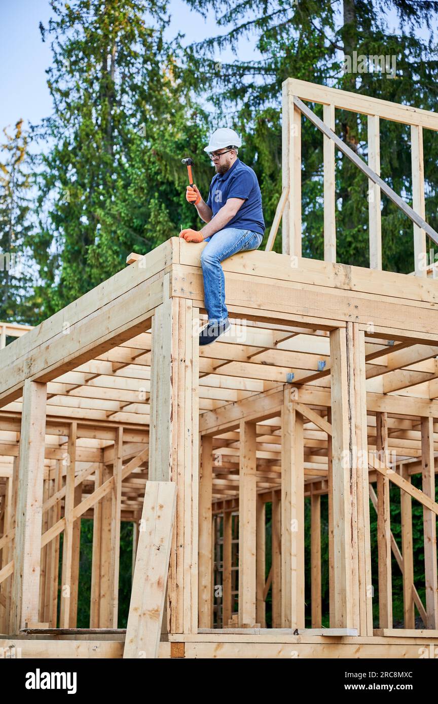 Joiner constructing wooden two-story frame house next to the forest. Bearded man in glasses ...