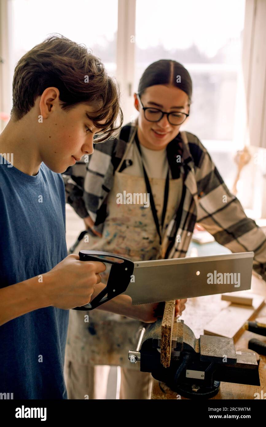 Male teenage student cutting wood while standing with female teacher ...