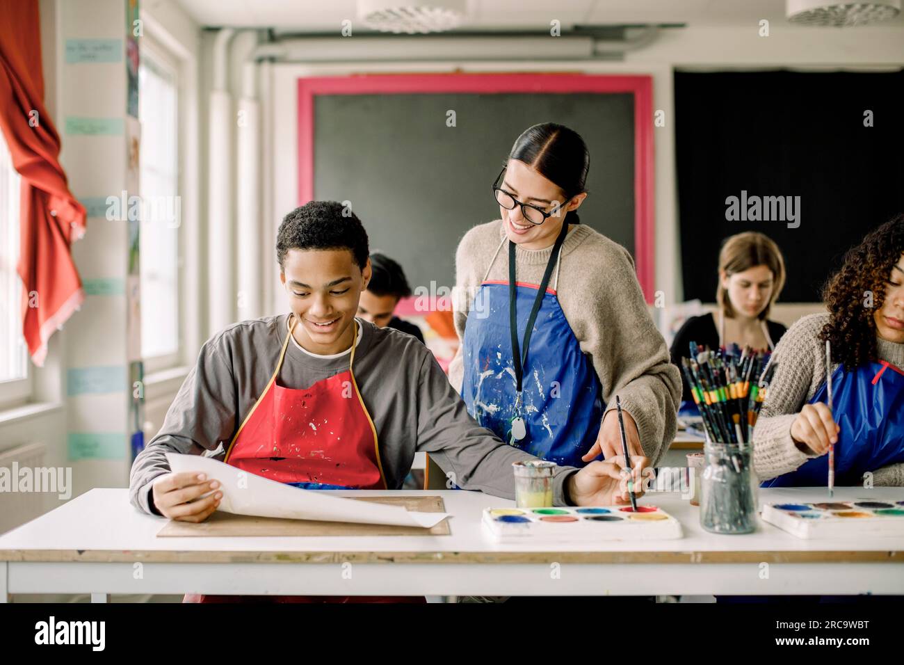 Smiling male teenage student showing artwork to female teacher during ...