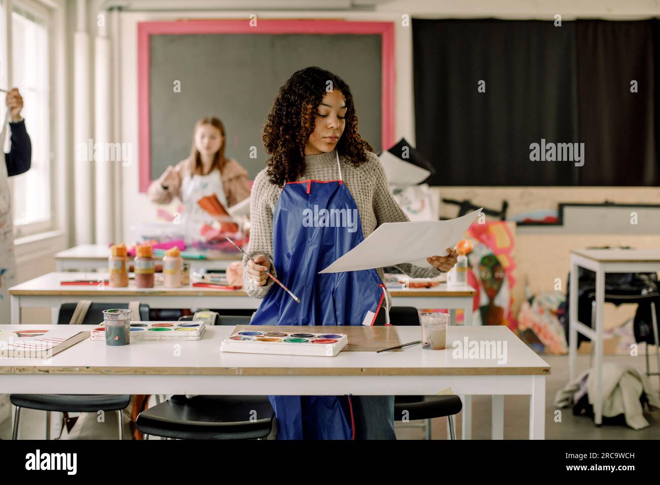 Teenage female student examining painting standing near desk during art ...