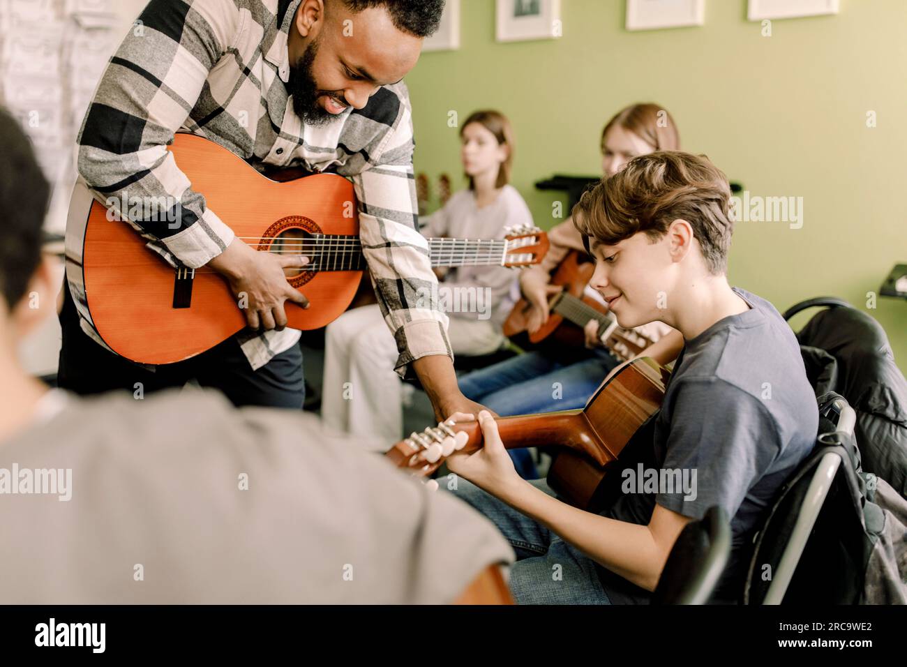 Young male teacher assisting teenage boy playing guitar in music class ...