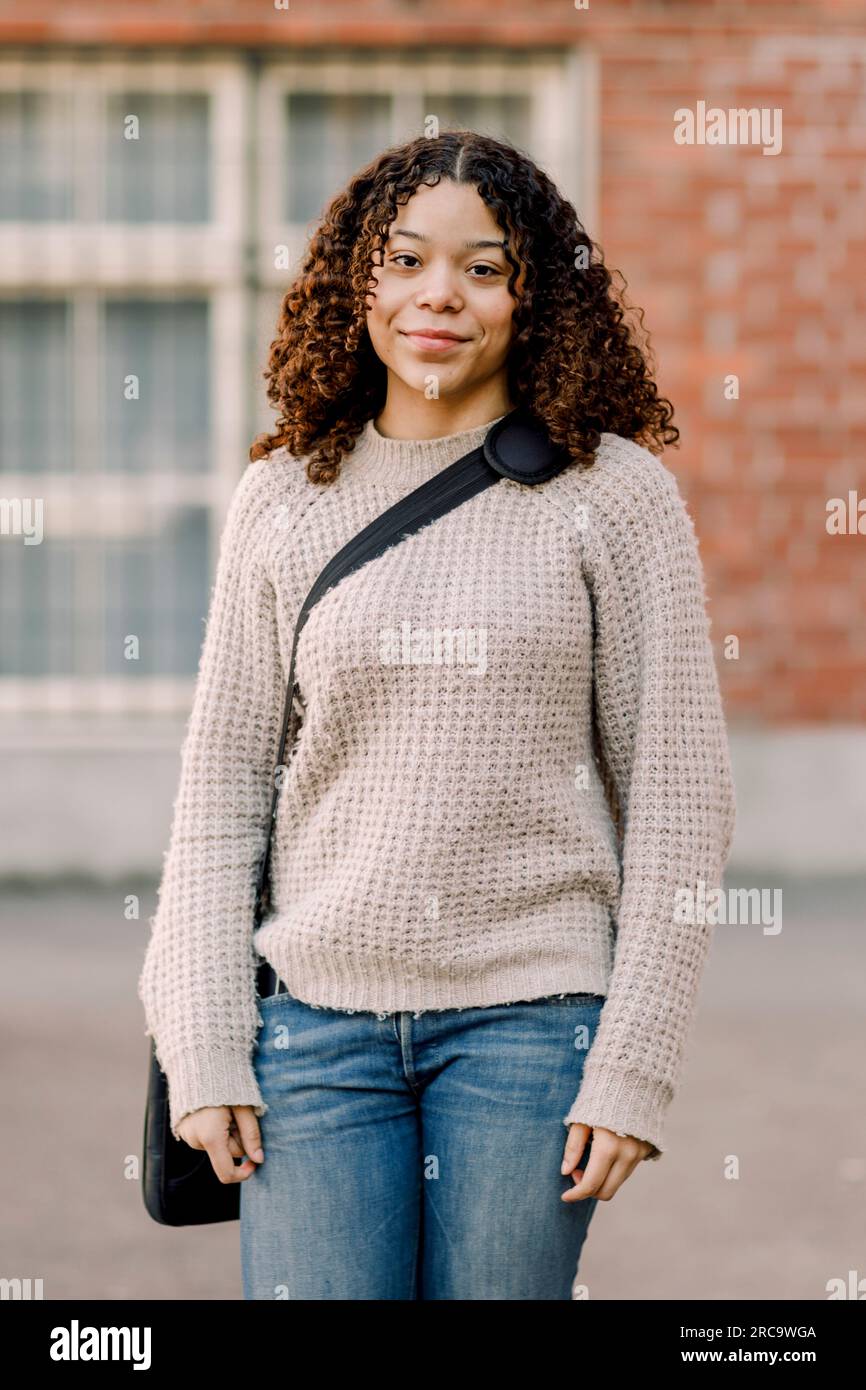 Portrait of smiling female teenage student with curly hair standing in ...