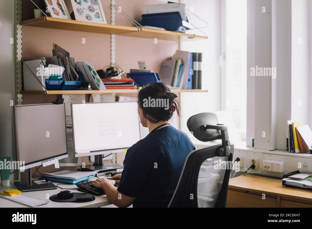 Rear view of mature female nurse working on computer in clinic Stock ...