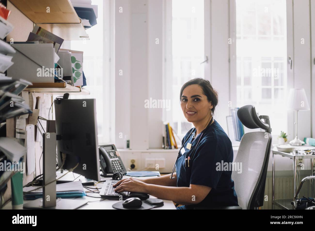 Side view portrait of female nurse sitting at desk with computer in ...