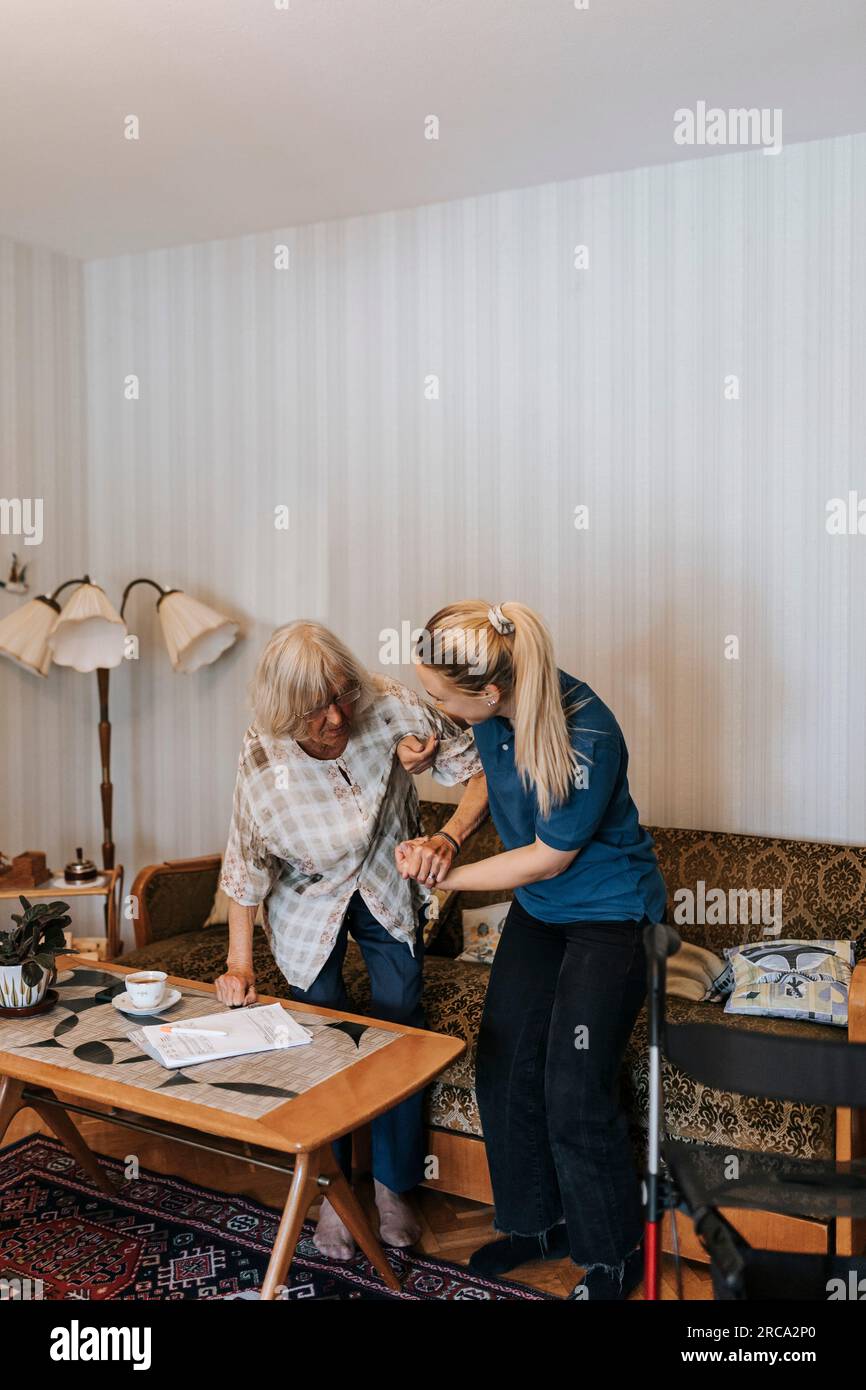 Female Caregiver Assisting Senior Woman While Standing Up At Home Stock