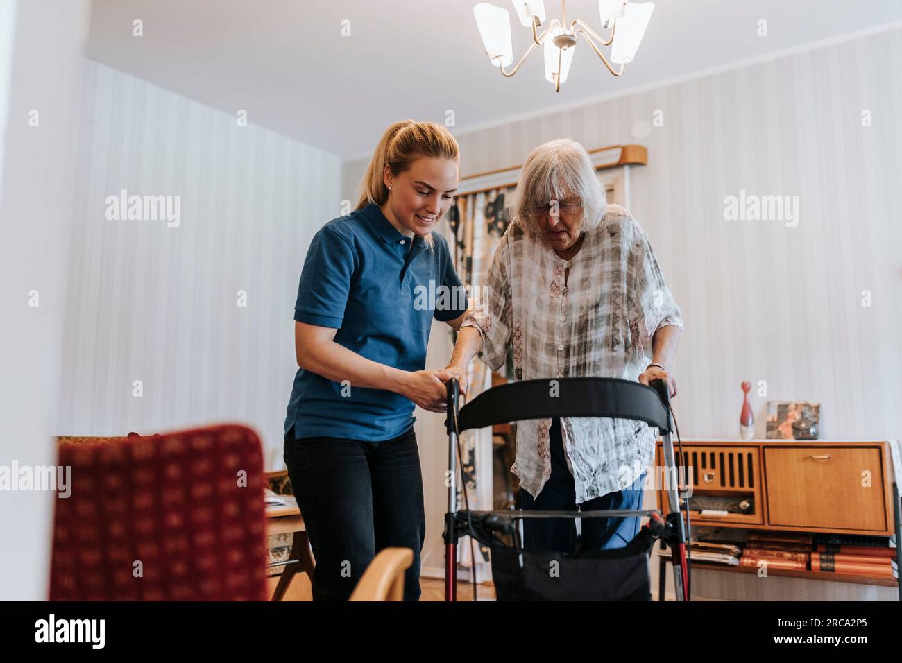 Female caregiver supporting senior woman using mobility walker at home ...
