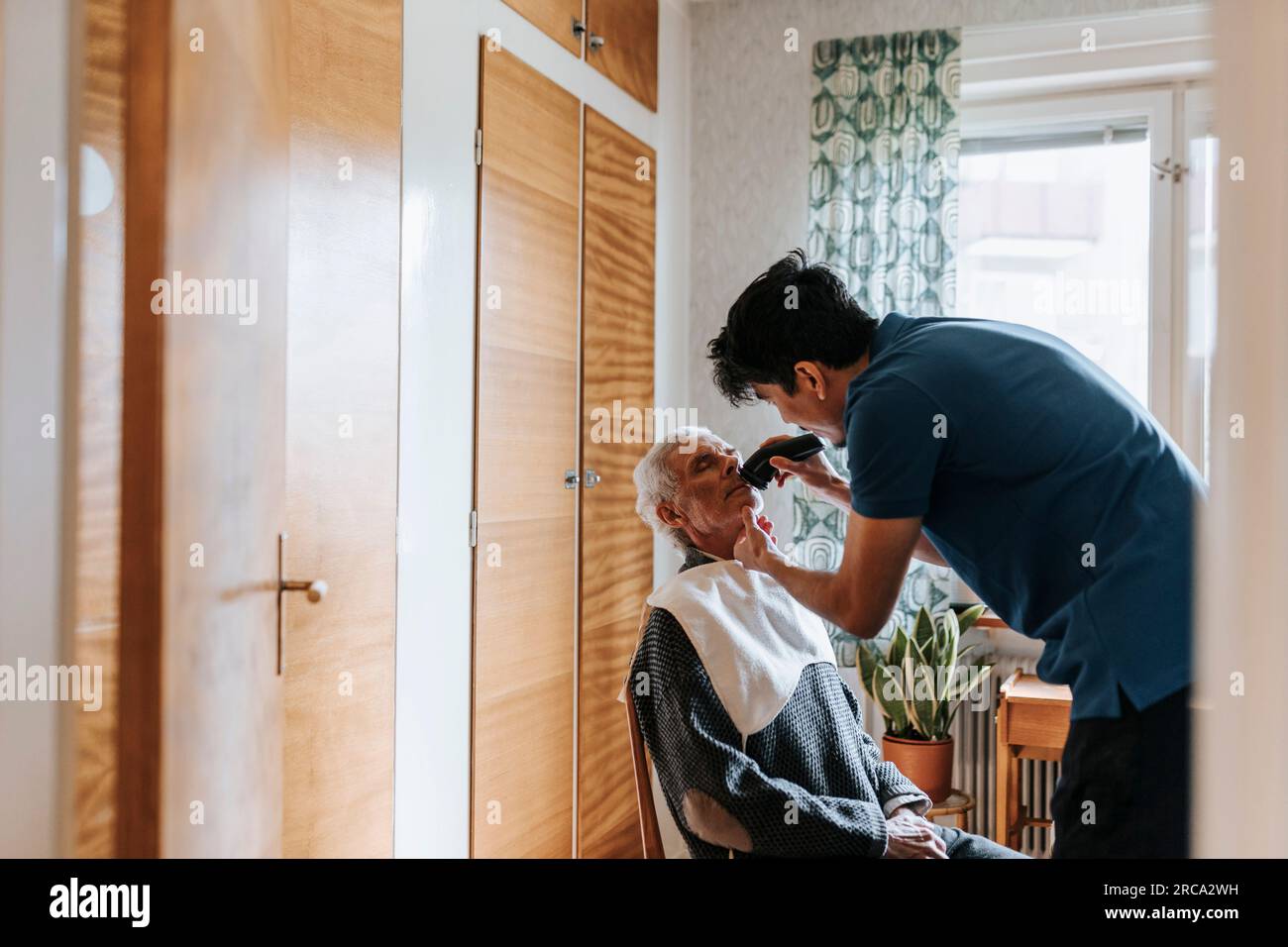 Side view of male care assistant trimming mustache of senior man at ...
