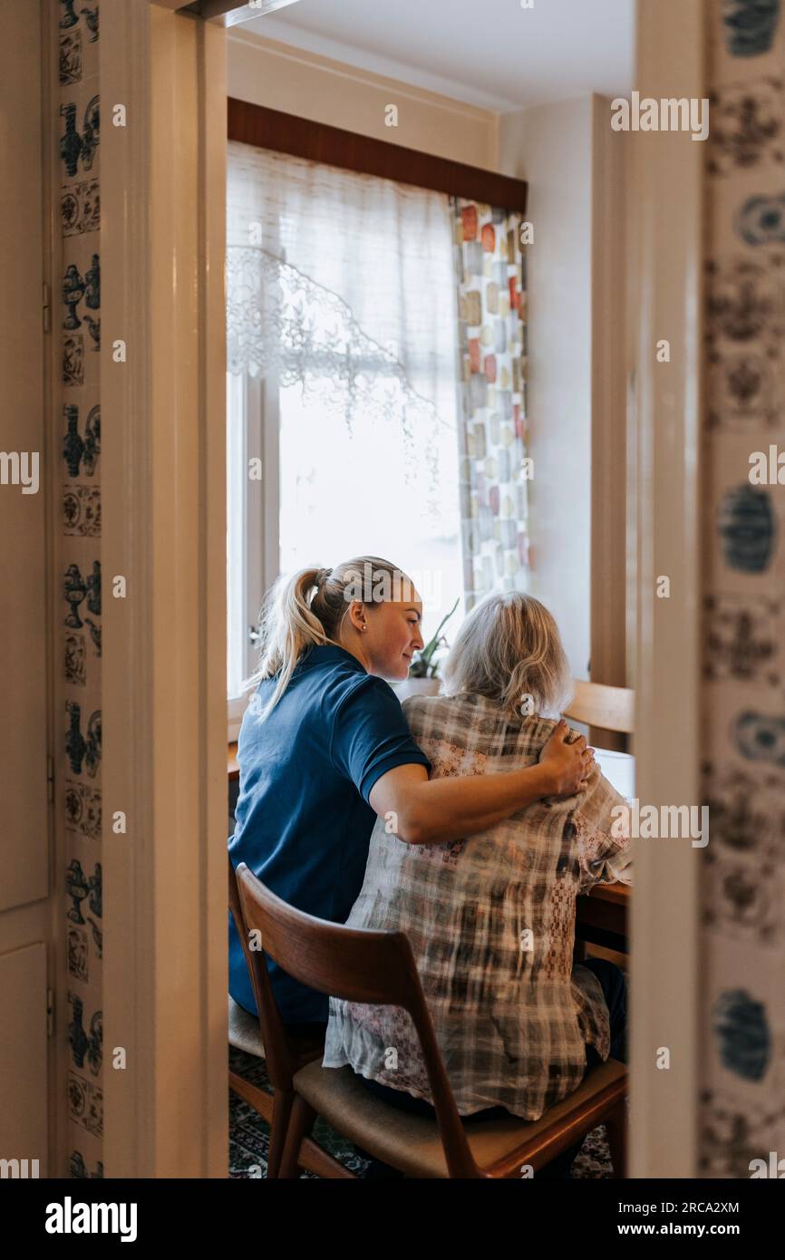 Female caregiver with arm around of senior woman sitting on chair at ...