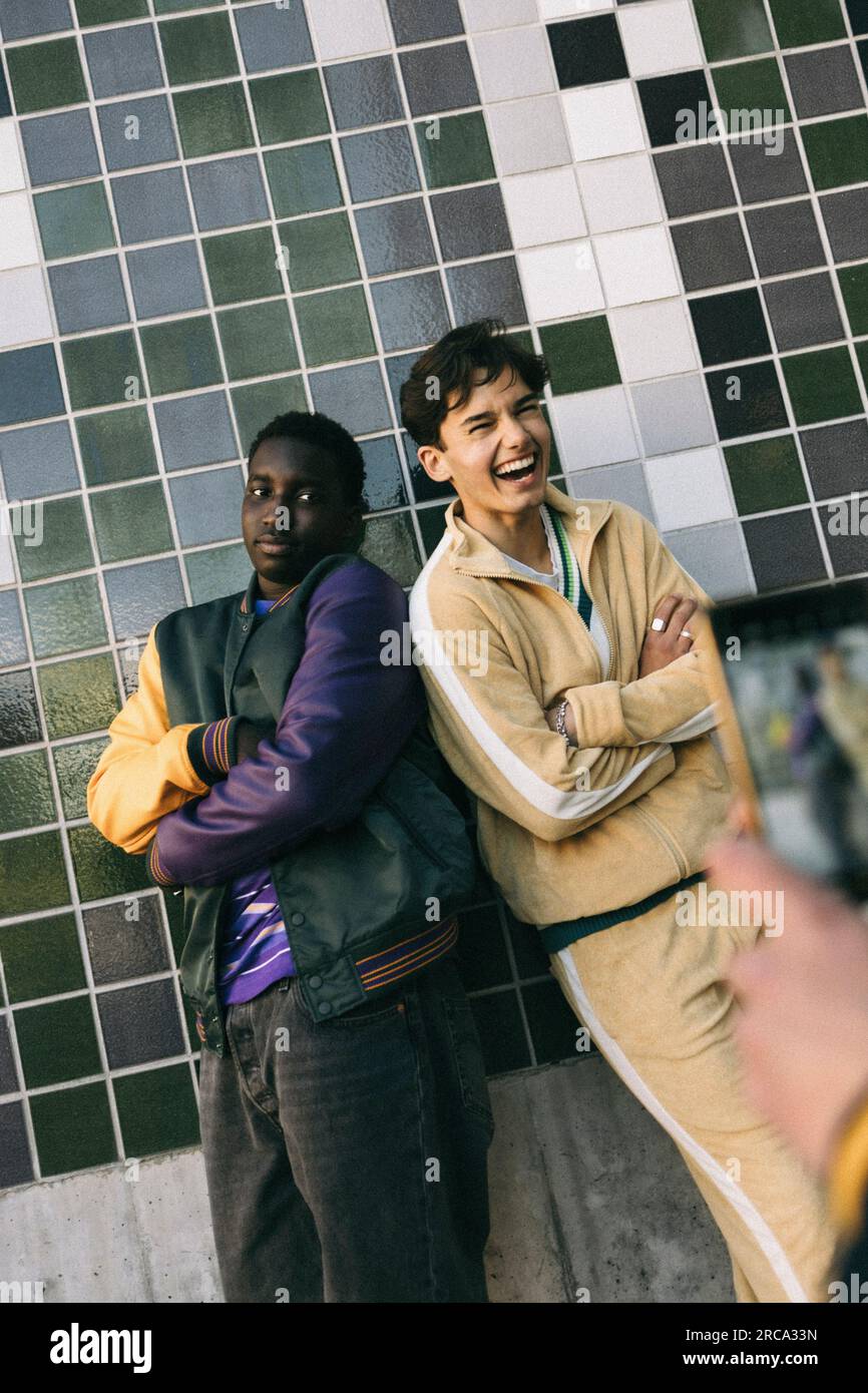 Young man laughing while standing with male friend against tiled wall ...