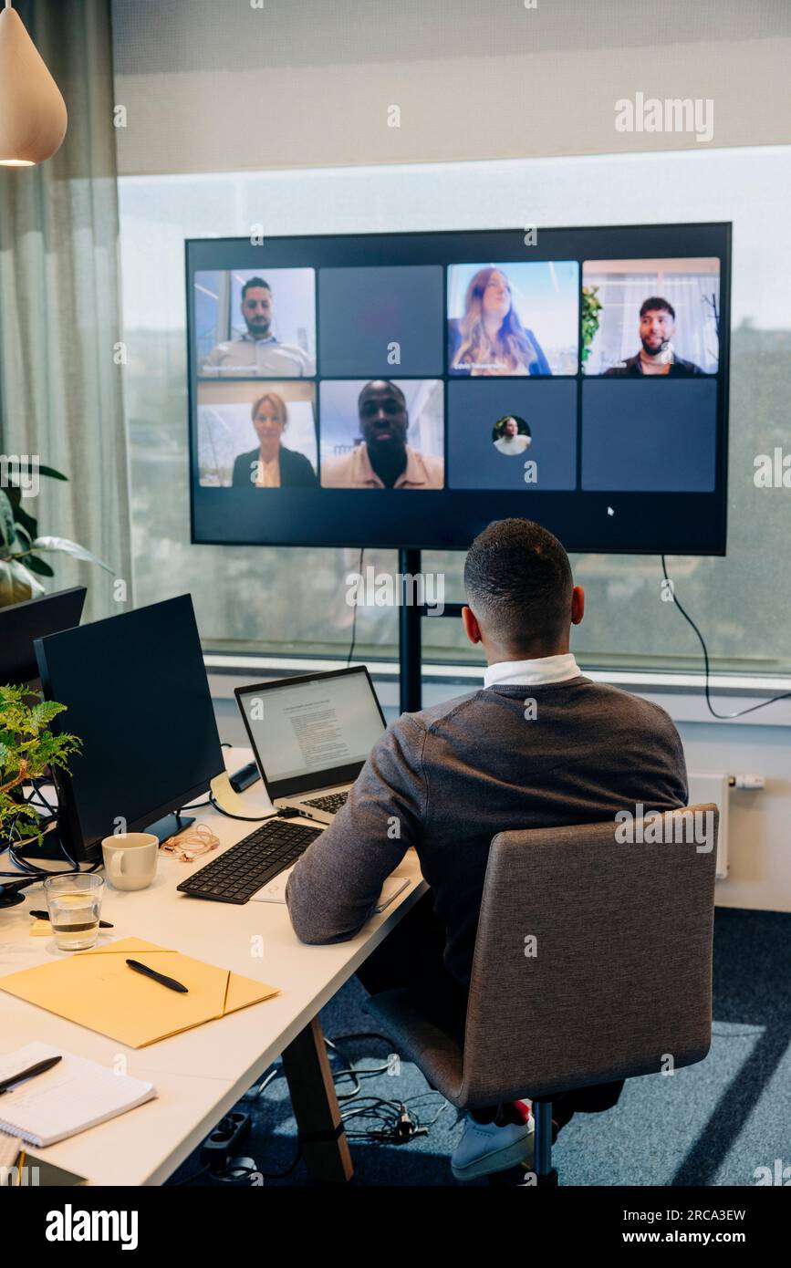 Rear view of businessman doing video conference with colleagues on TV ...