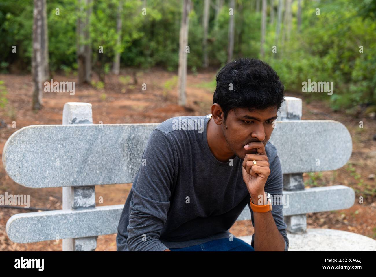 A sad Indian man is sitting on a bench in a park. He is looking down at ...