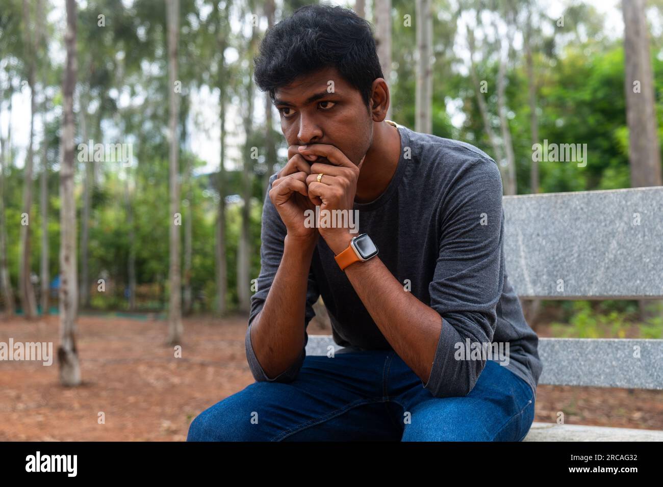A sad Indian man is sitting on a bench in a park. He is looking down at ...