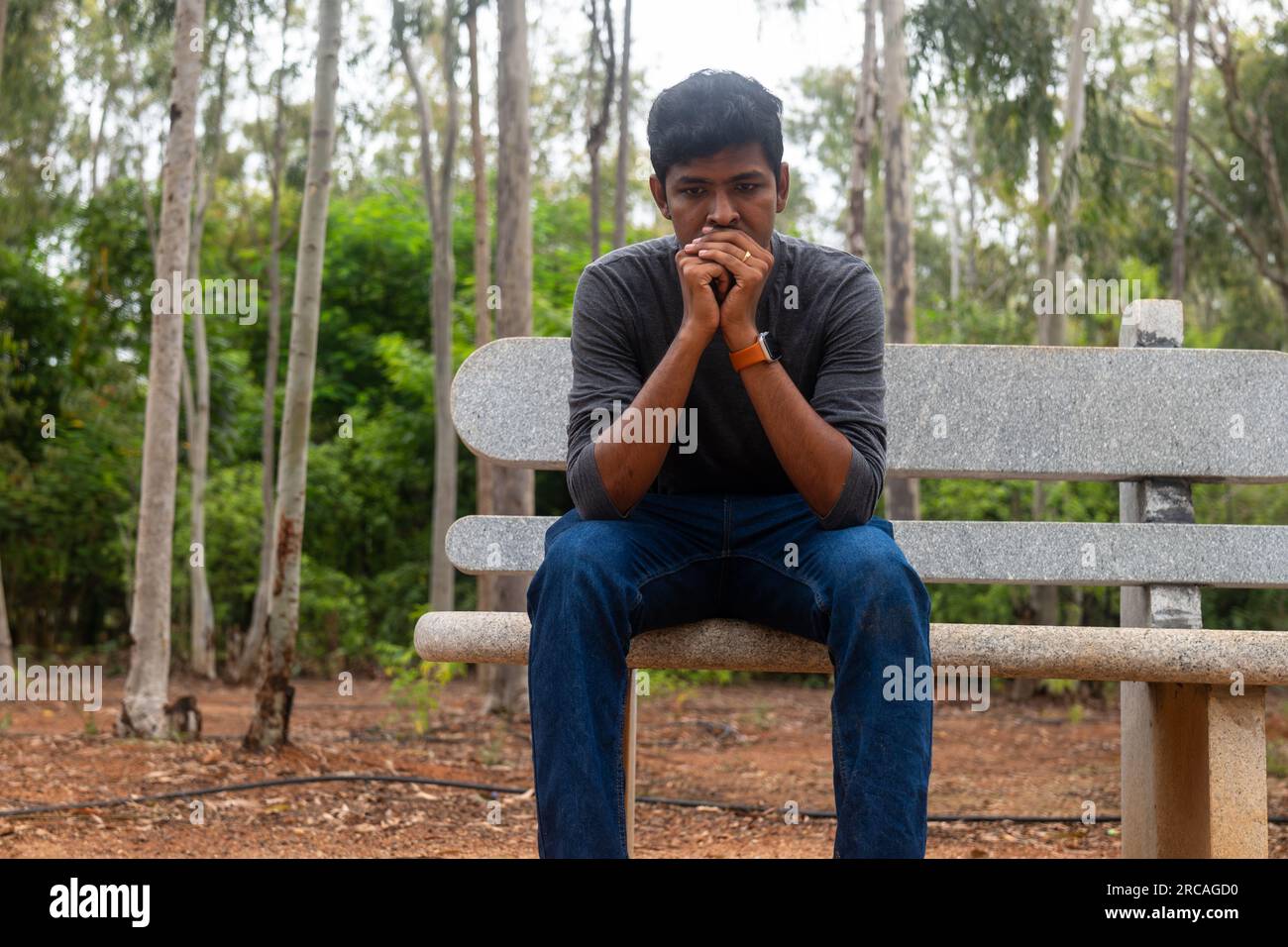 sad Indian man sitting on a bench in a park. He is looking down at his ...