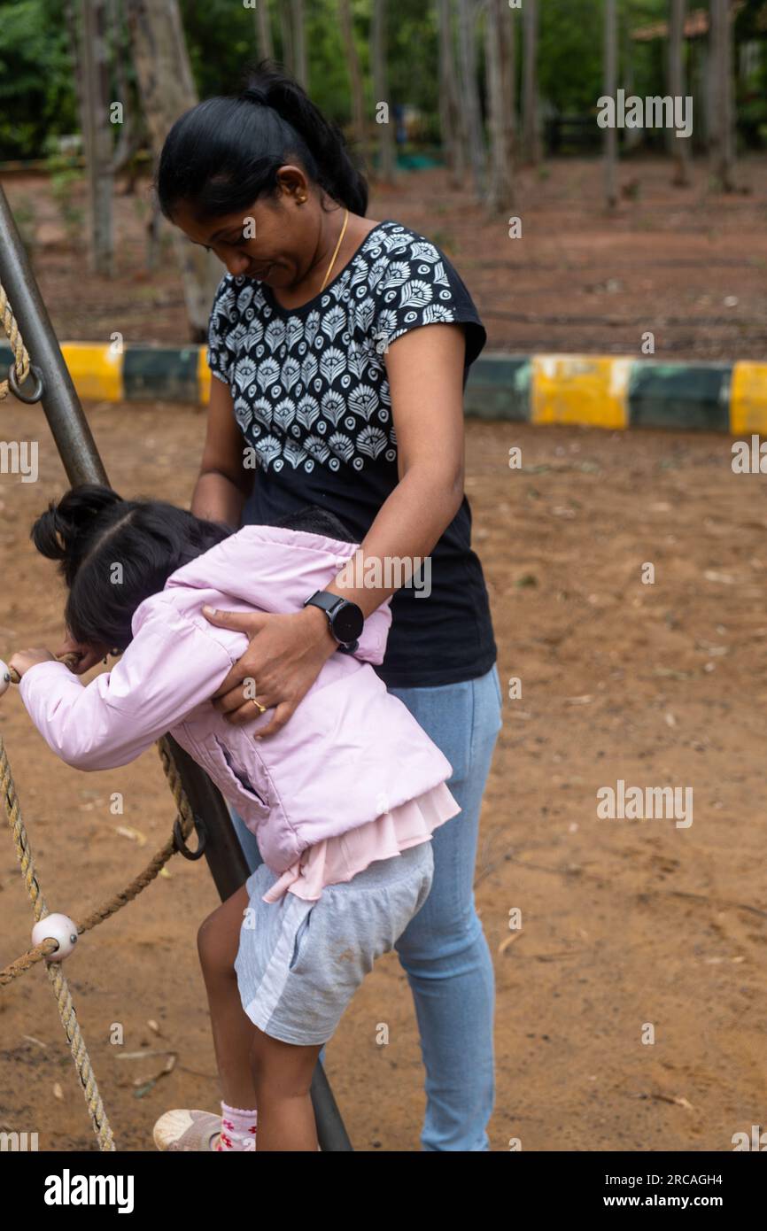 A mother helps her young daughter climb a rope in a park. The mother is ...