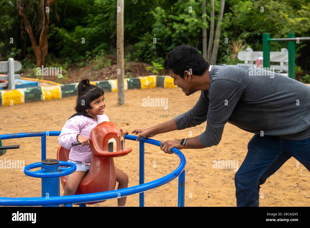 A father and daughter twirl on a spinner in a park. The father is ...