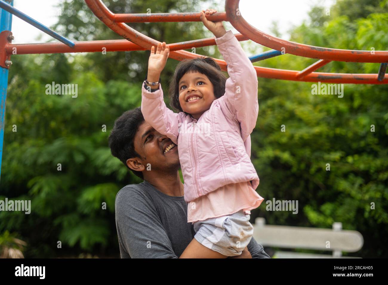 A father and daughter are climbing monkey bars in a park. The father is ...