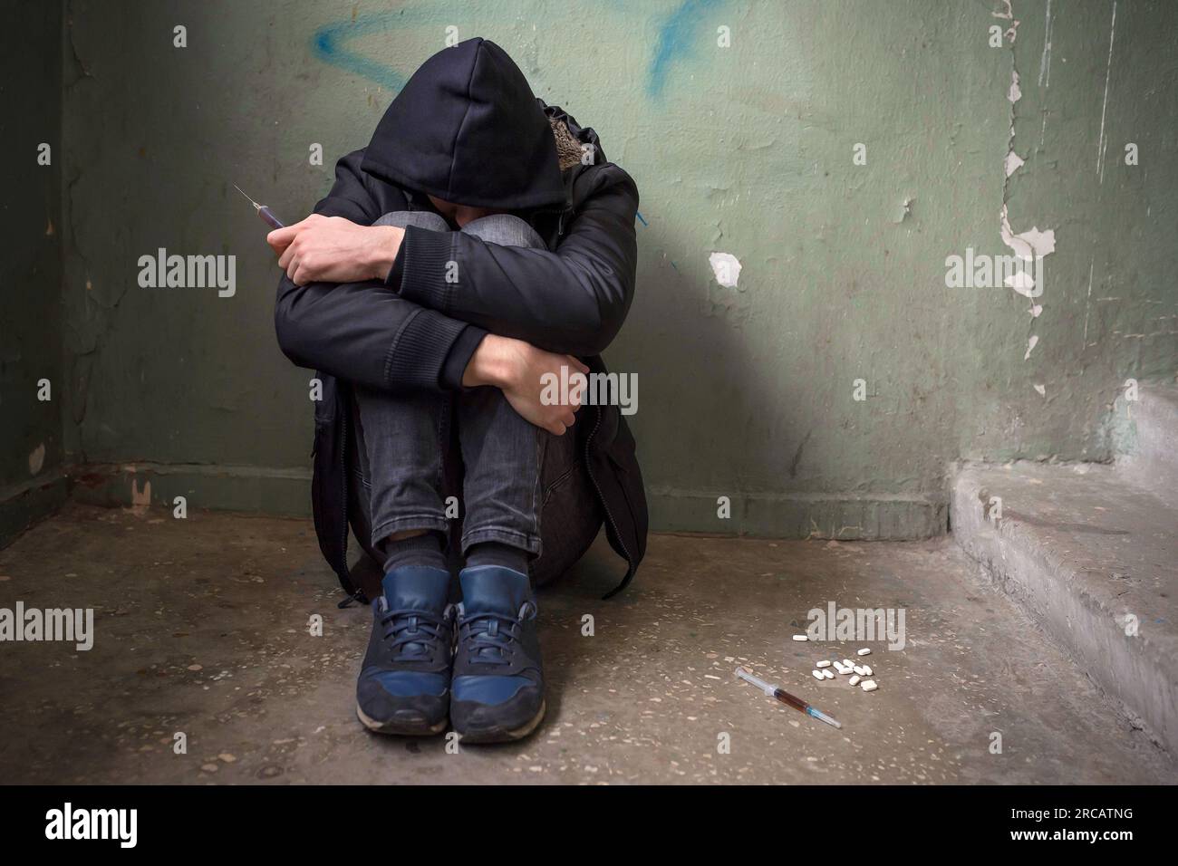 Addict with a syringe in the hood and sitting on the floor in an ...