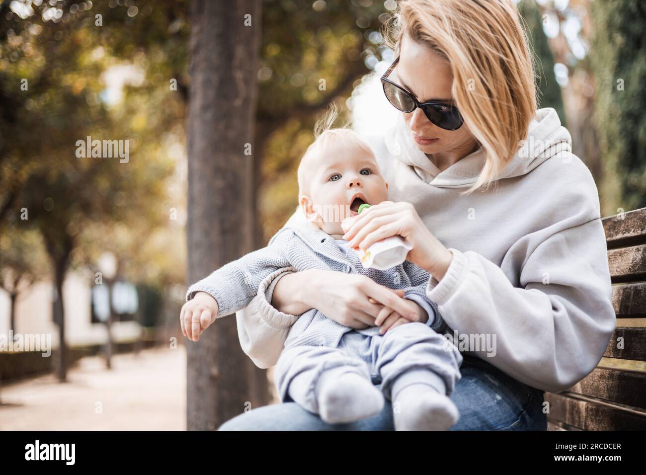 Mother sitting on bench in urban park, holding her infant baby boy ...