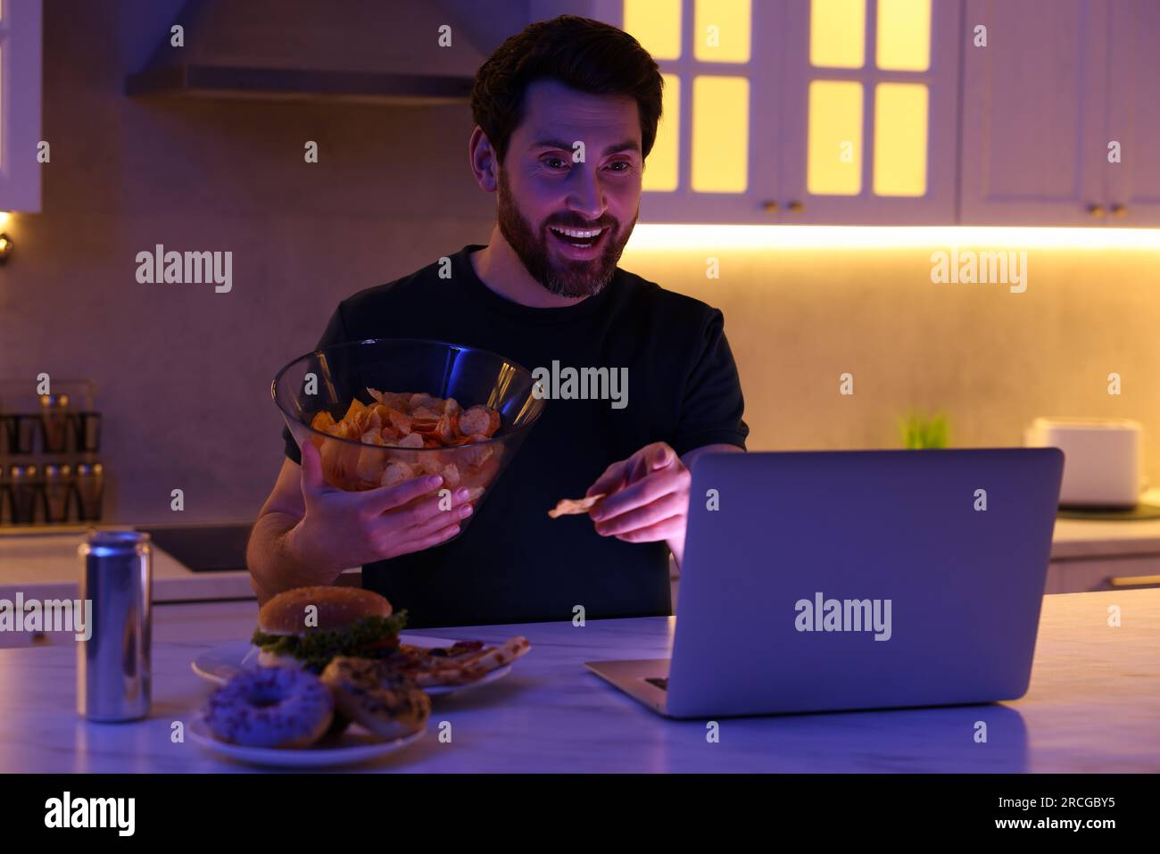 Man eating chips while using laptop in kitchen at night. Bad habit ...