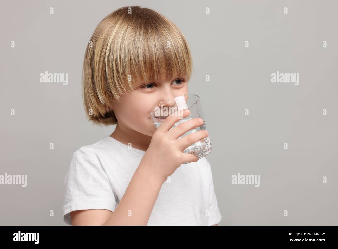 Cute little boy drinking fresh water from glass on light grey ...
