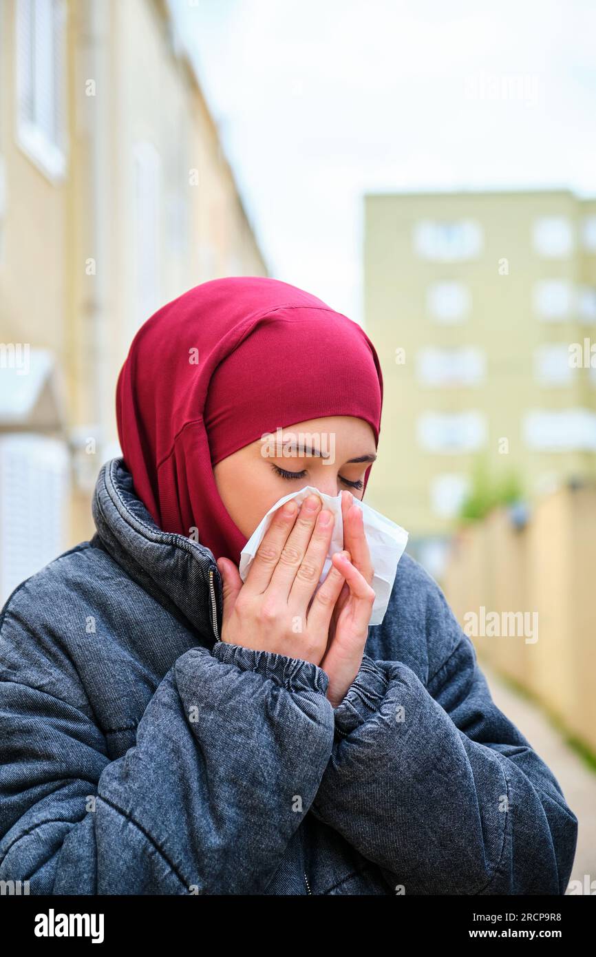 Muslim young woman in hijab and coat blowing her nose in a tissue in ...