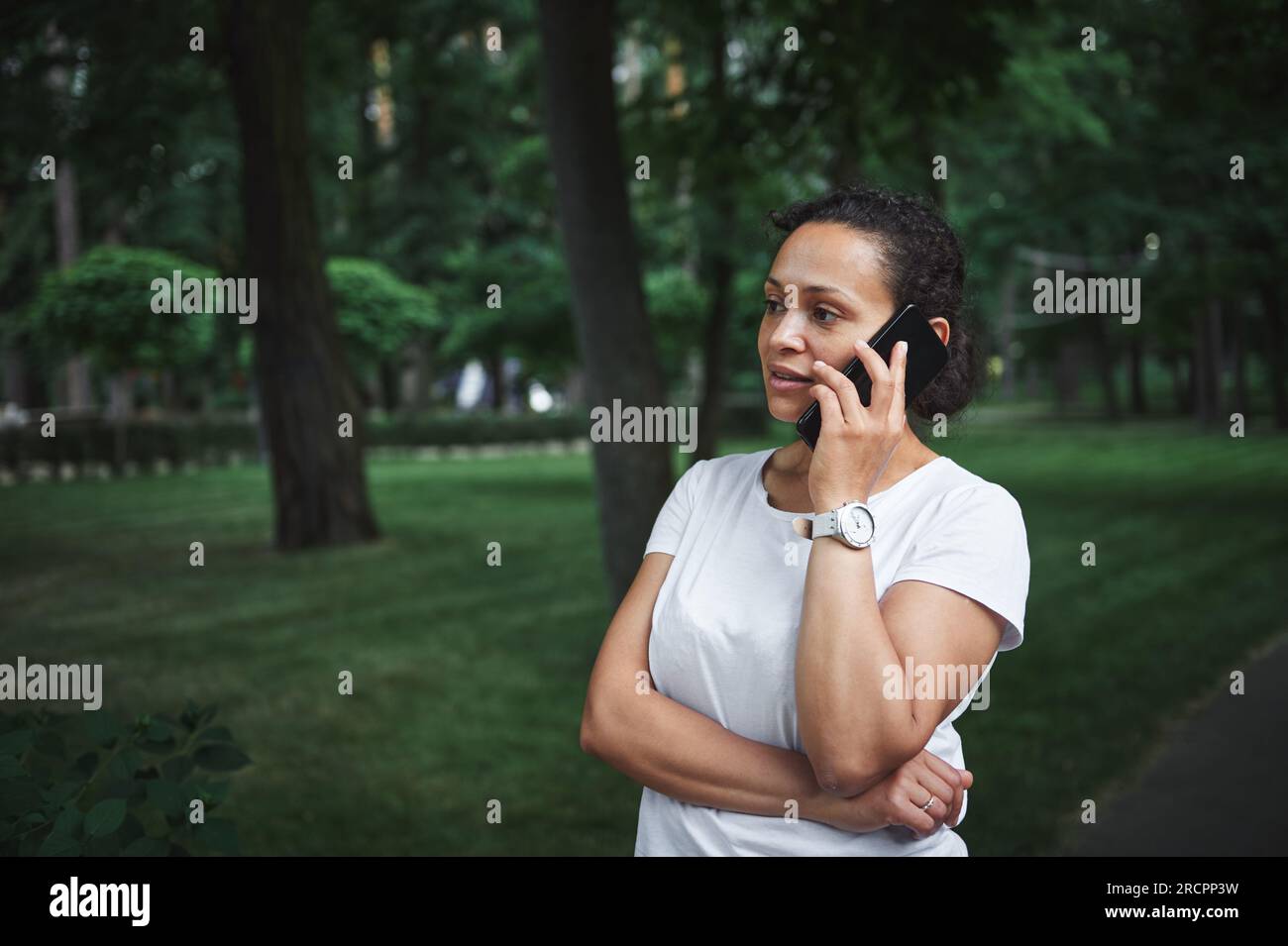 Confident multi-ethnic young pretty woman in white t-shirt, smiling, talks on cell phone while ...