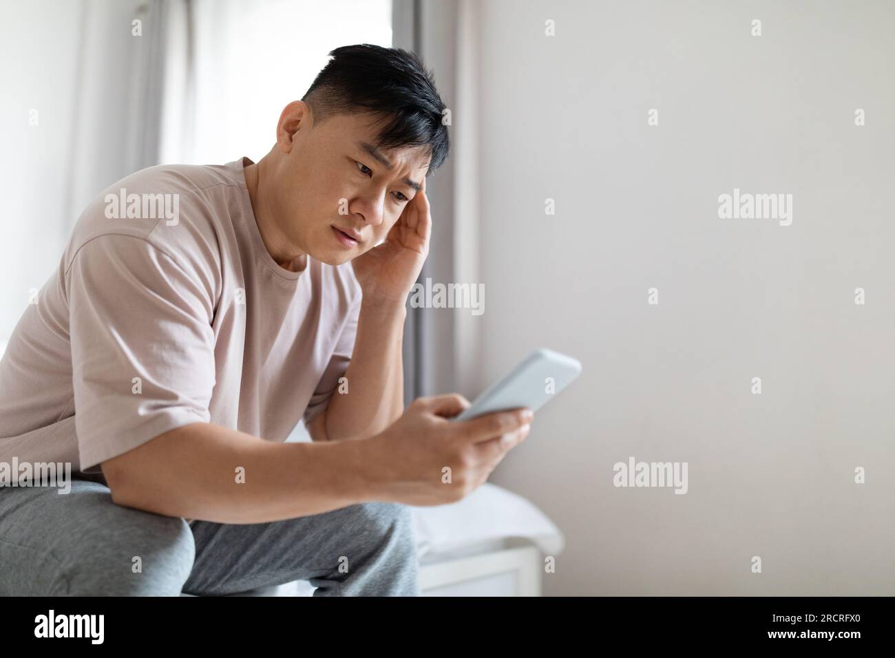 Worried upset asian man sitting on bed, using smartphone Stock Photo ...