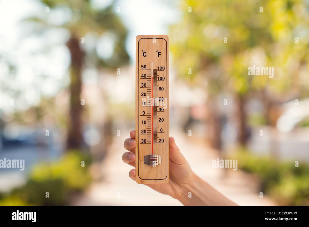 Hot weather. Thermometer in hand in front of street with trees and ...