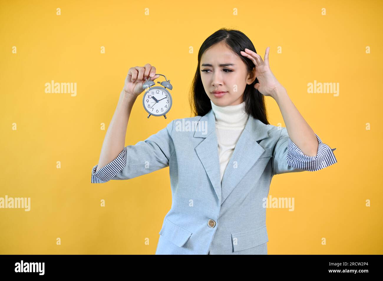 A perplexed Asian businesswoman, looking at an alarm clock in her hand ...