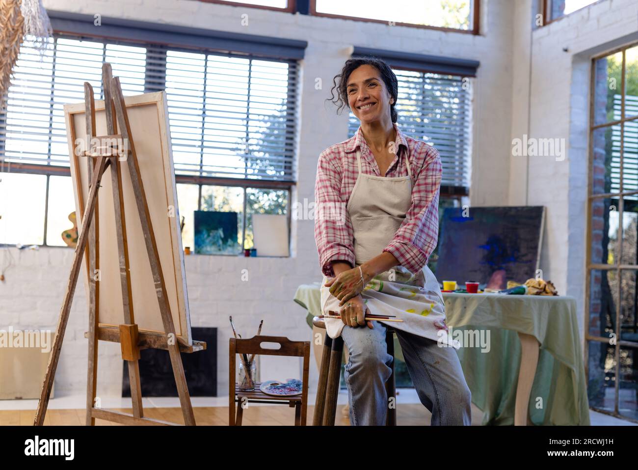 Happy biracial woman wearing apron holding brushes in home studio Stock ...