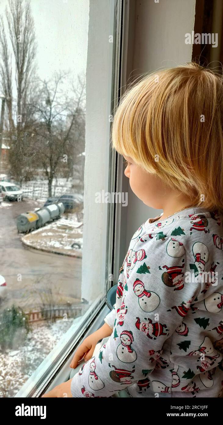A little boy with blonde hair sits by the window on the windowsill and ...