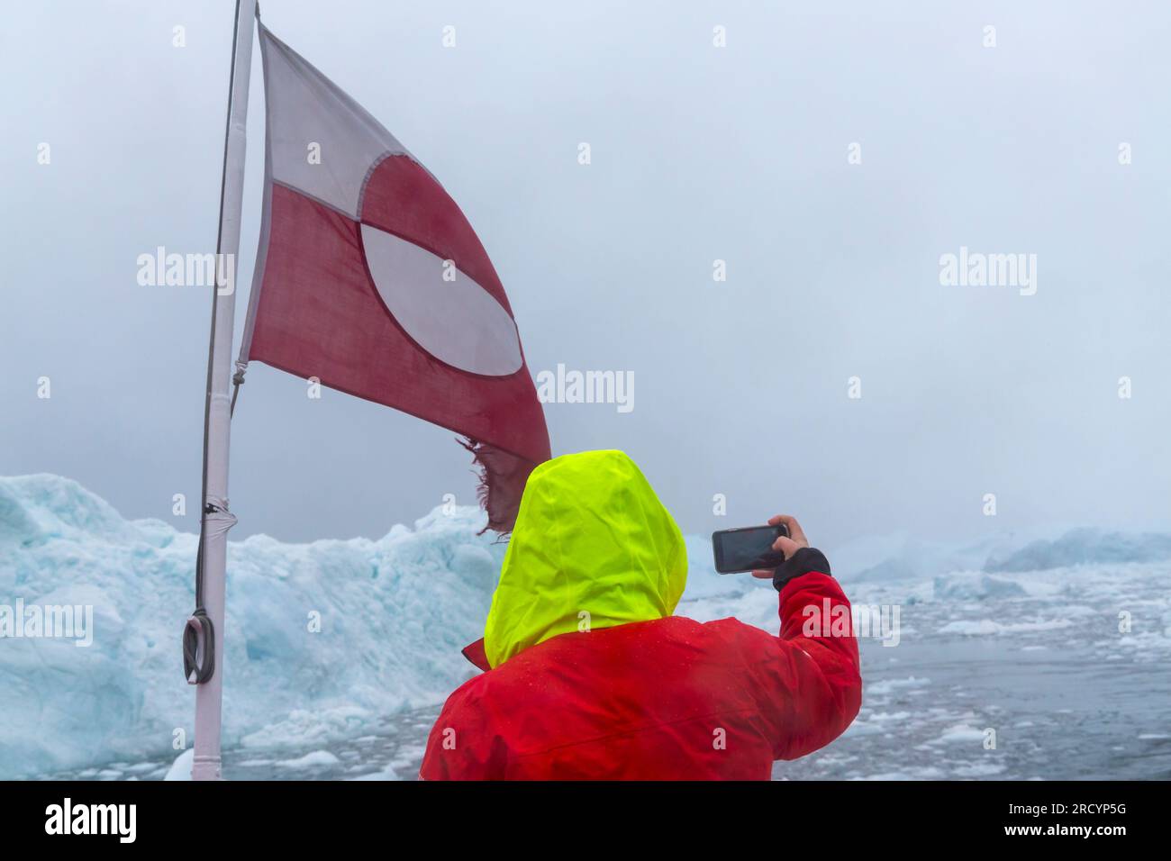 Tourist taking a photo on smartphone by Greenland flag with iceberg ...